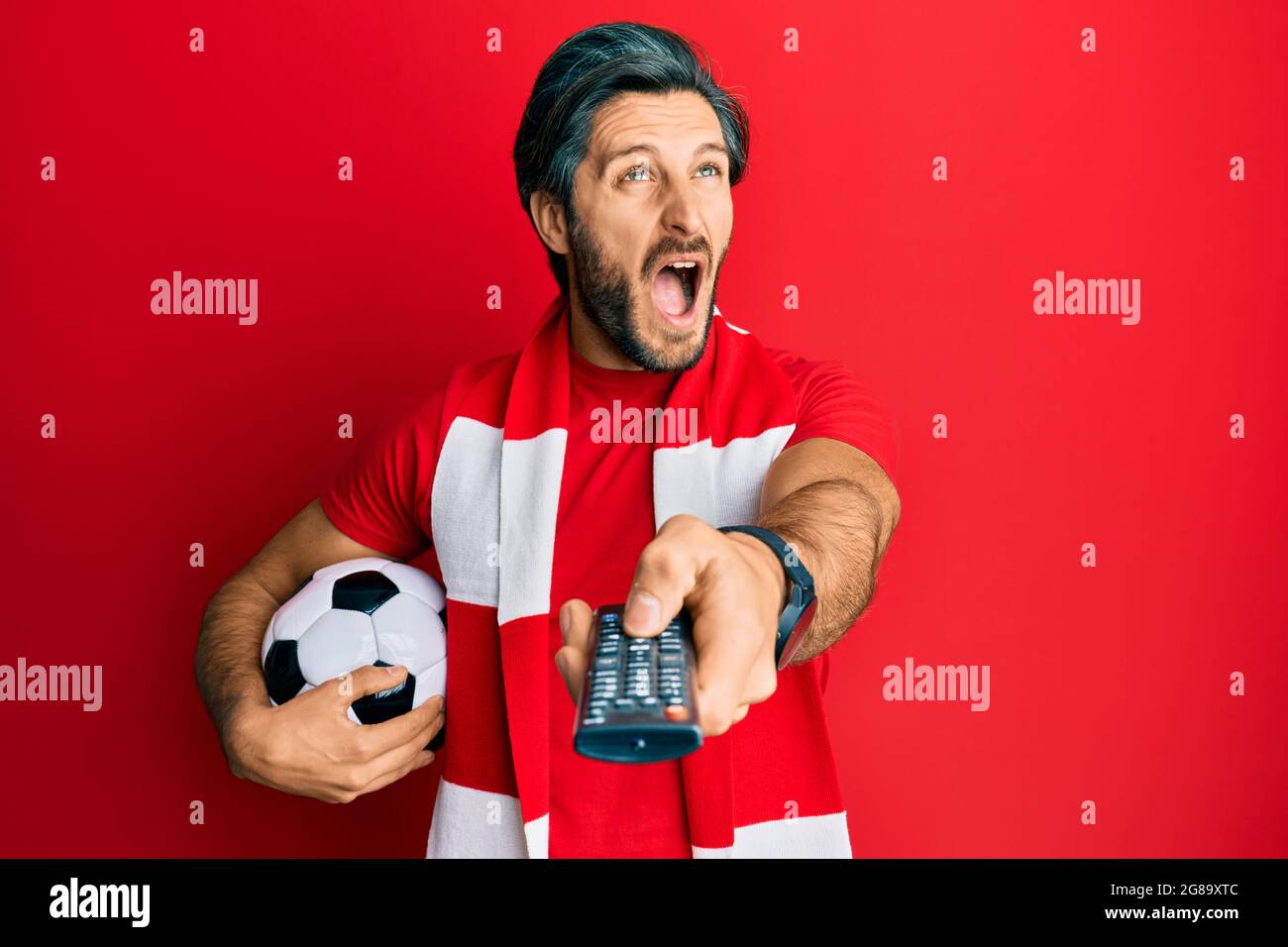 Young hispanic man football hooligan holding ball using tv control ...