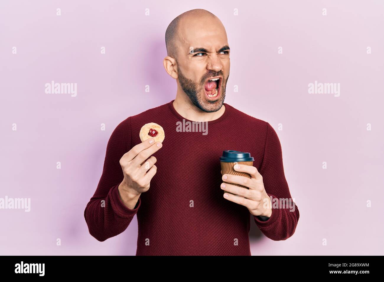 Young bald man drinking coffee and eating pastry angry and mad ...