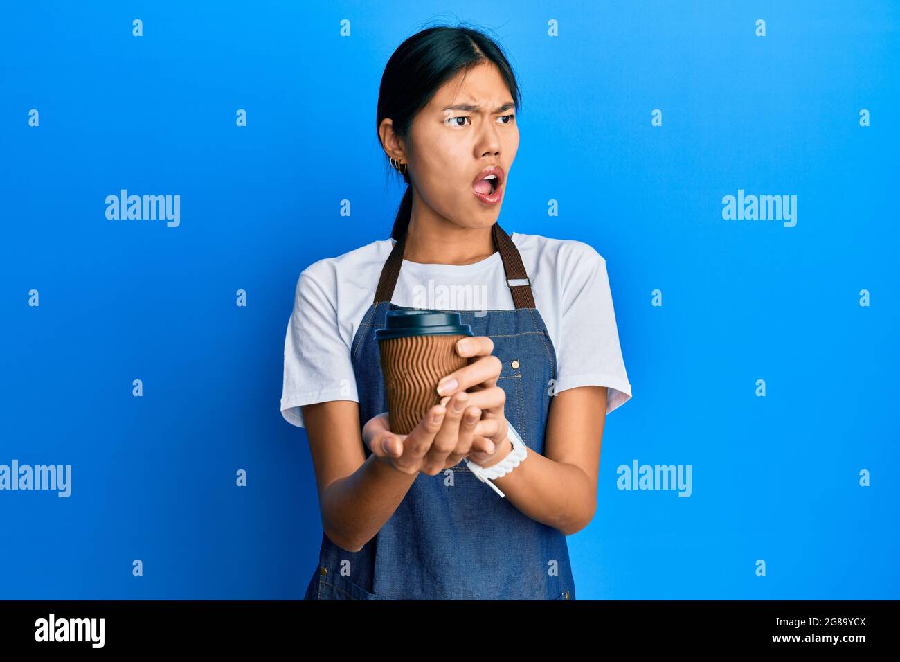 Young chinese woman wearing waiter apron holding cup of coffee angry ...
