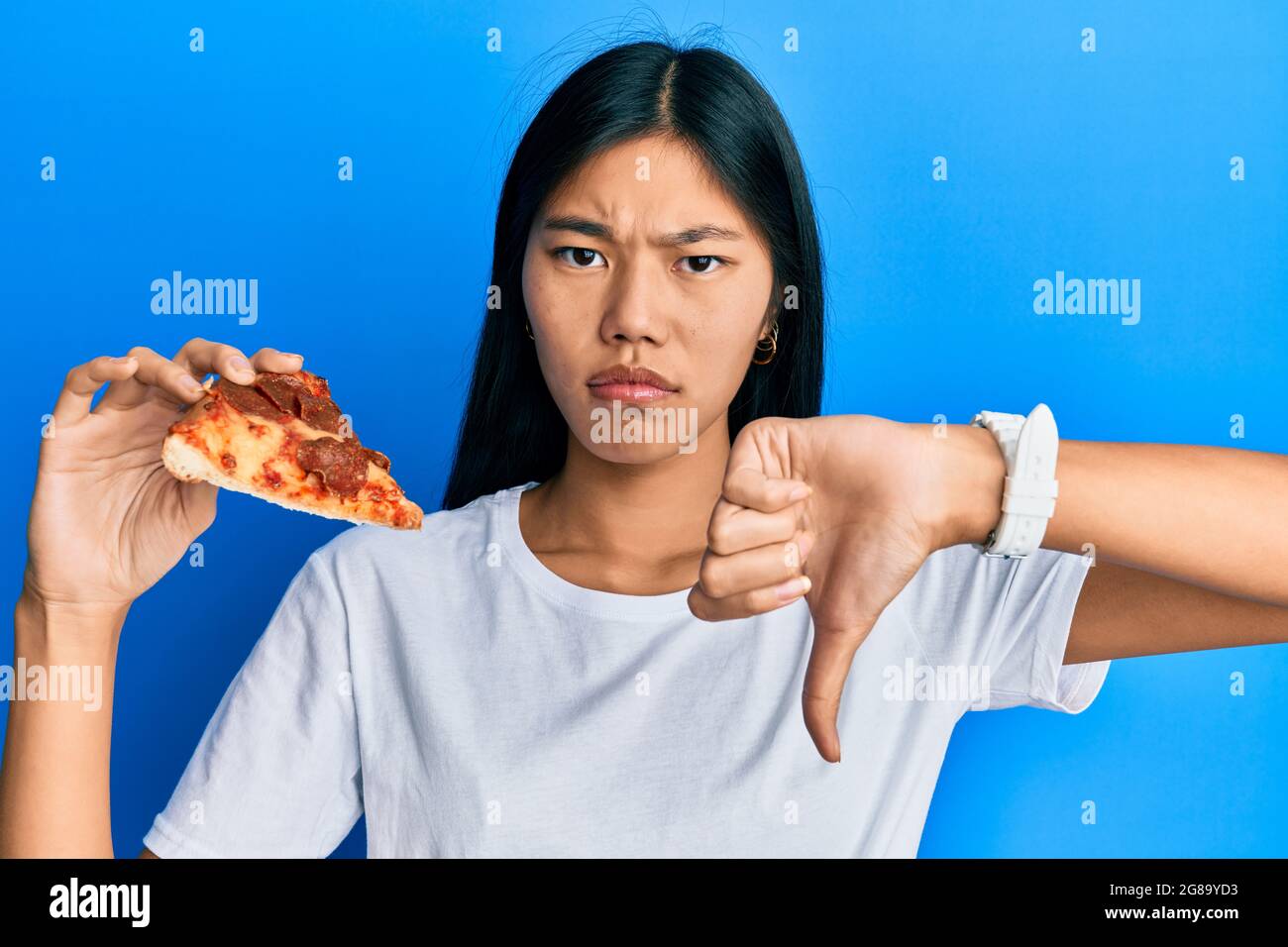 Young chinese woman eating tasty pepperoni pizza with angry face ...