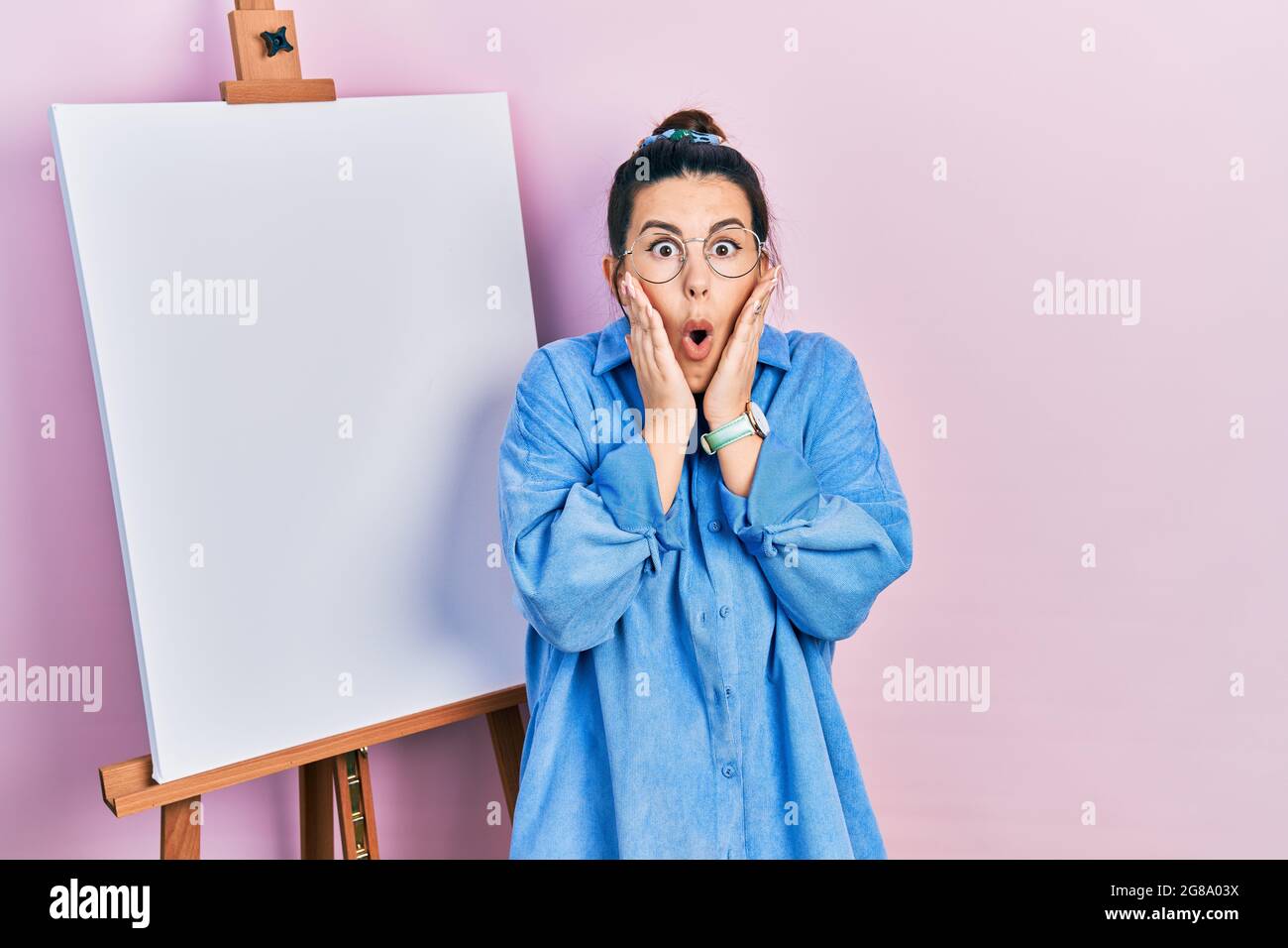 Young hispanic woman standing by painter easel stand afraid and shocked ...