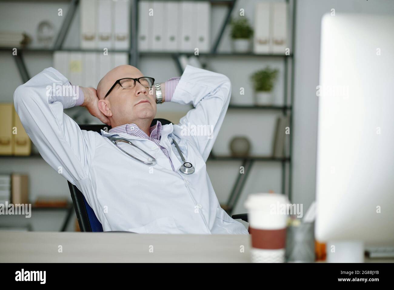 Mature doctor in white coat sitting at his workplace and relaxing after ...