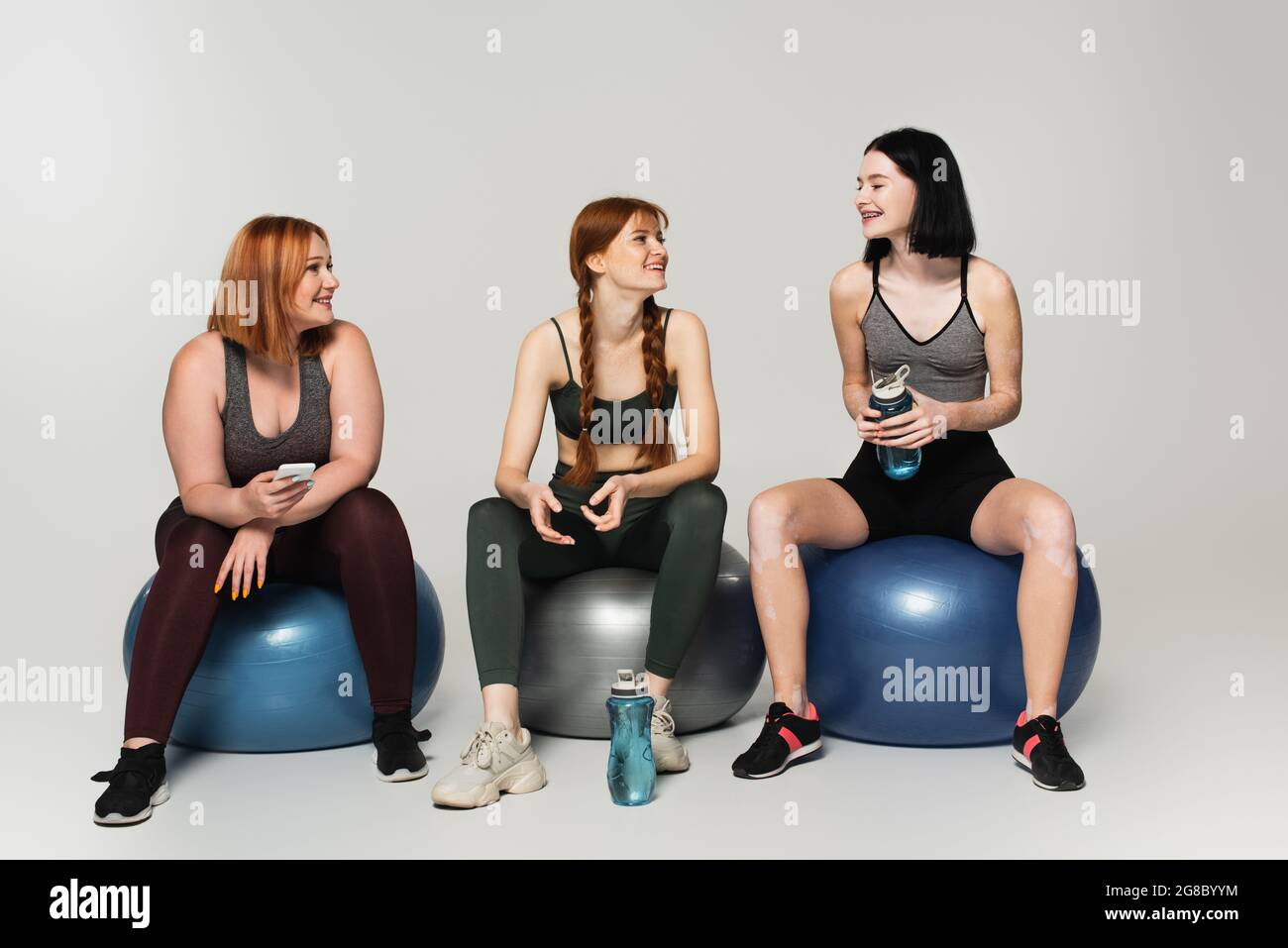 Body positive sportswoman with sports bottle looking at friends on ...