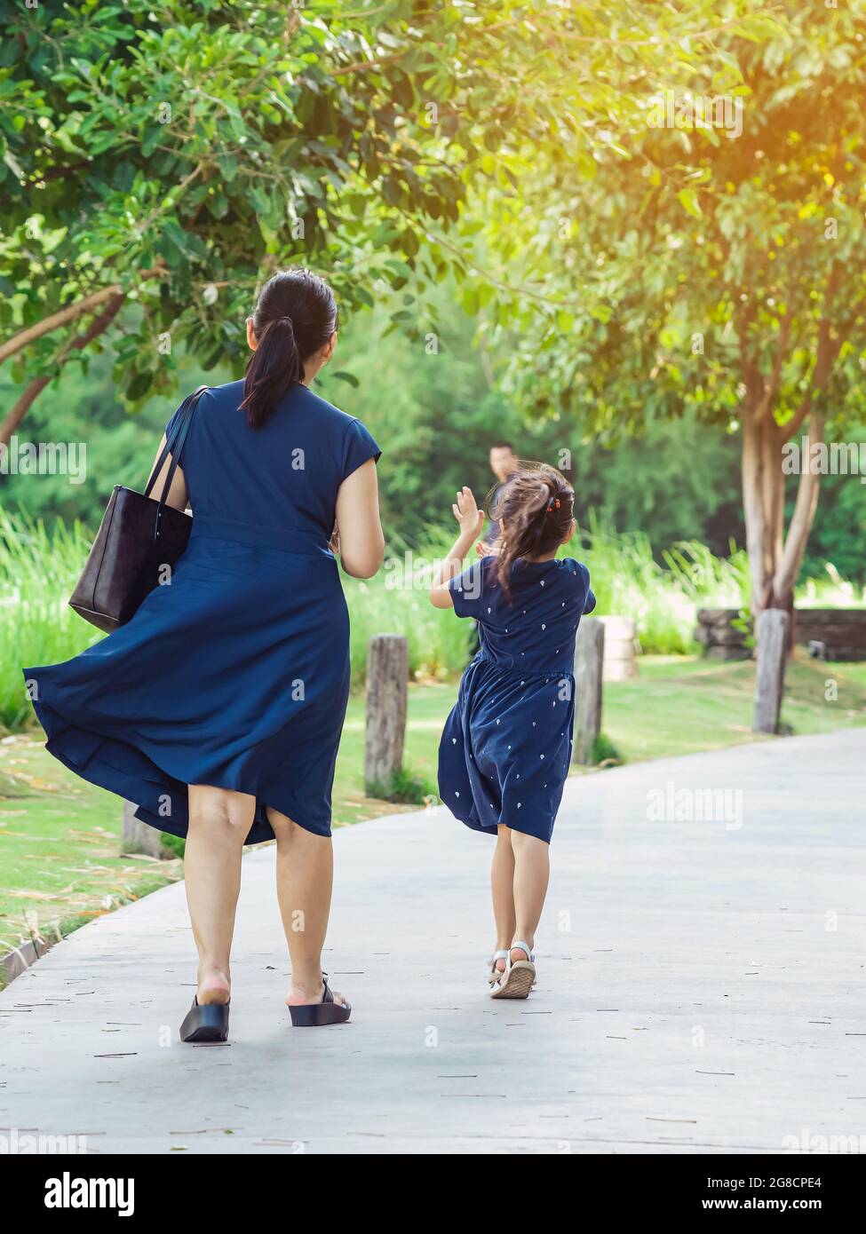 Back view of mother with lovely daughter in blue dress walking on pathway through green garden ...
