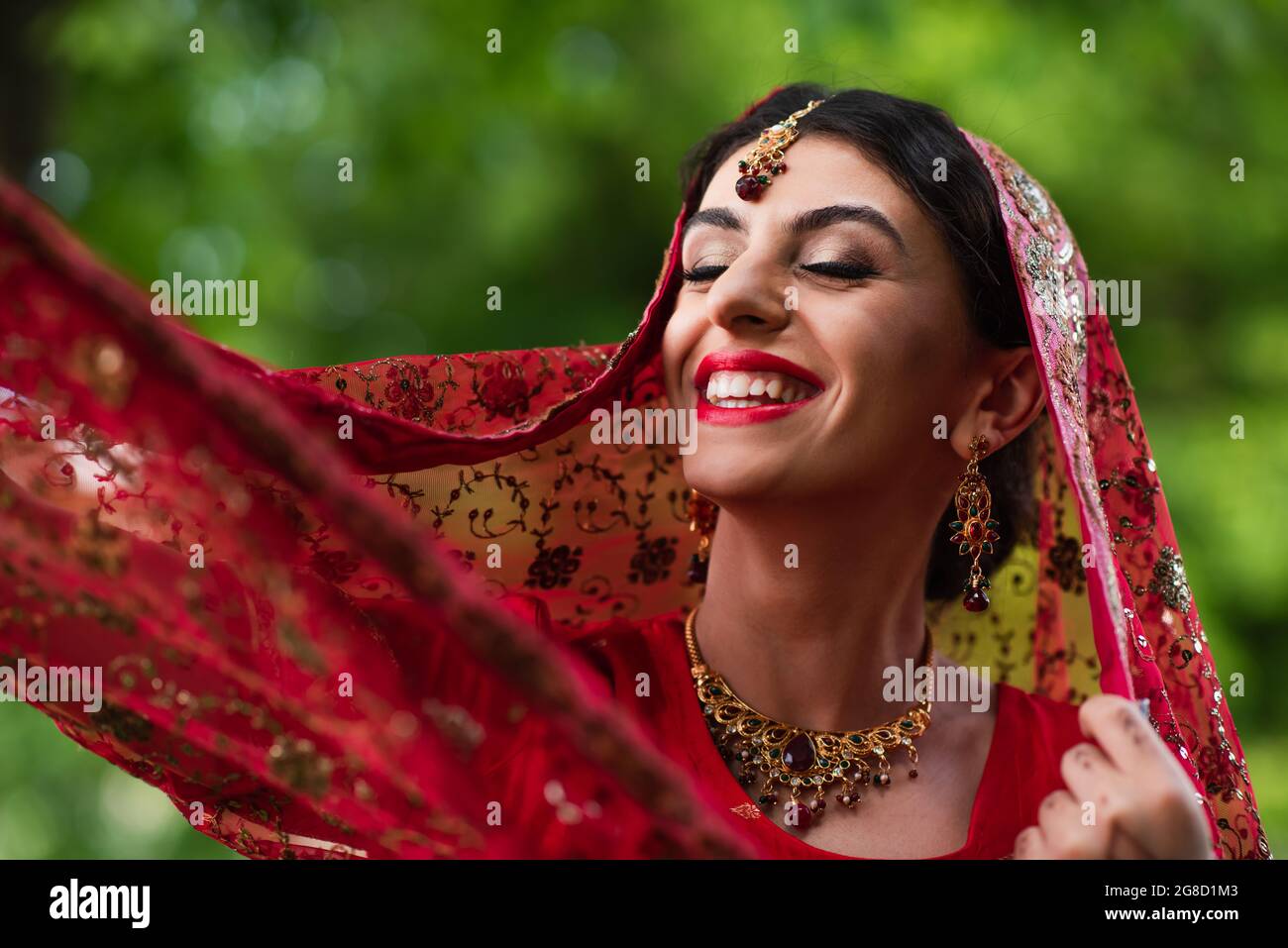 positive indian bride in red sari adjusting headscarf with ornament ...
