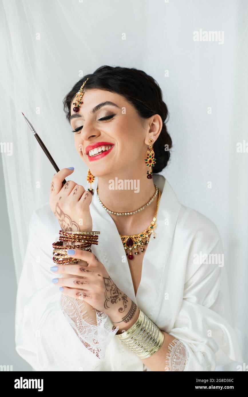 pleased indian bride with mehndi holding cosmetic brush on white Stock ...