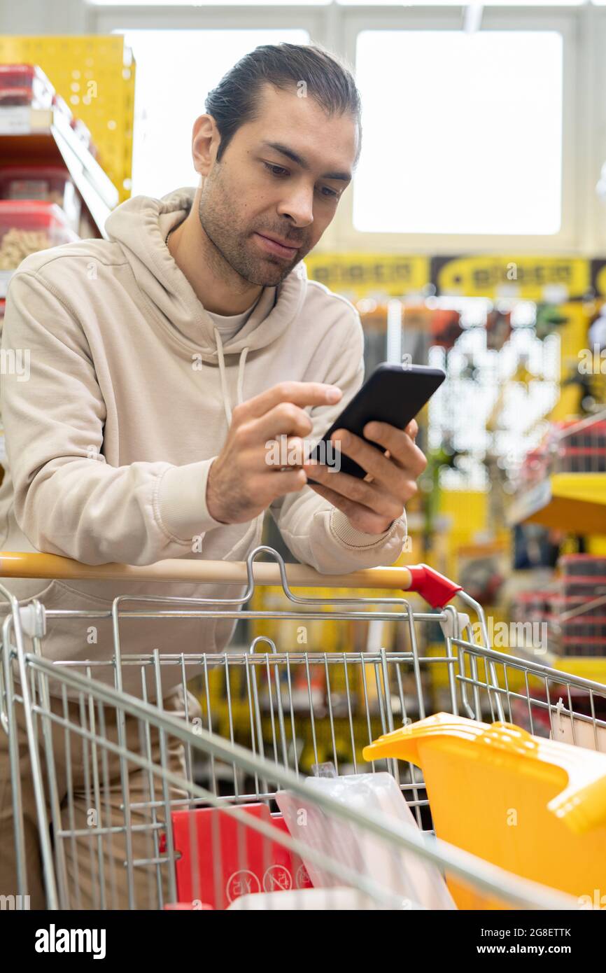 Male consumer with smartphone and shopping cart full of household goods ...