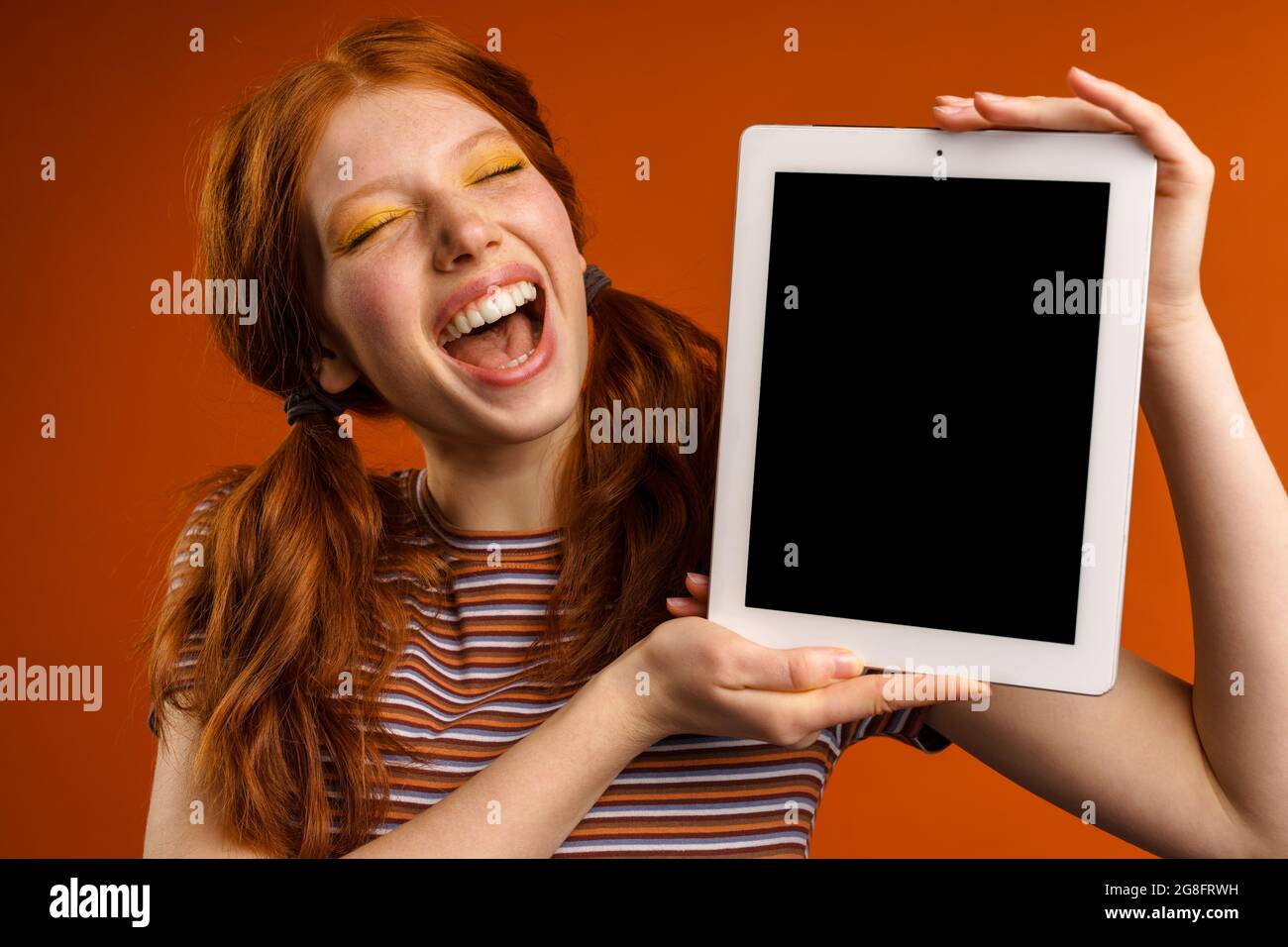 Ginger laughing woman with tails showing tablet pc isolated over orange ...