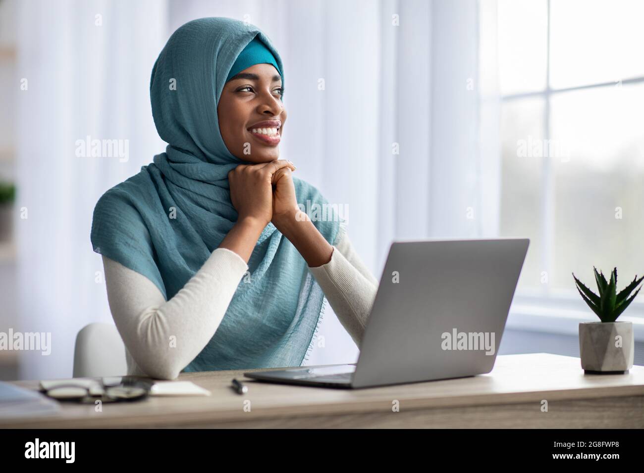 Pensive Happy Black Muslim Woman In Hijab Sitting At Desk With Laptop ...