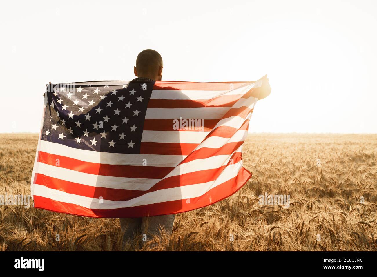 Young man holding American flag on back while standing in wheat field ...