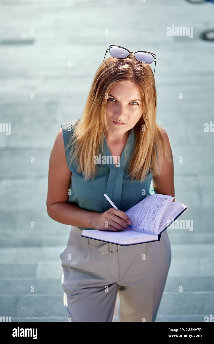 Strict young woman with sketchpad writes something outdoors using pen ...