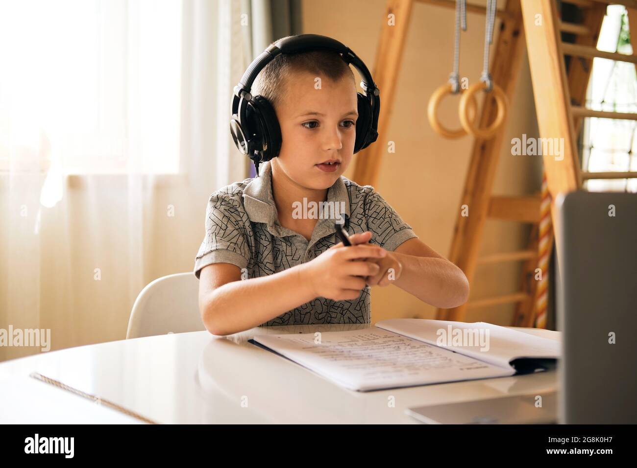 Young boy in headphones writing in notebook and using laptop while ...