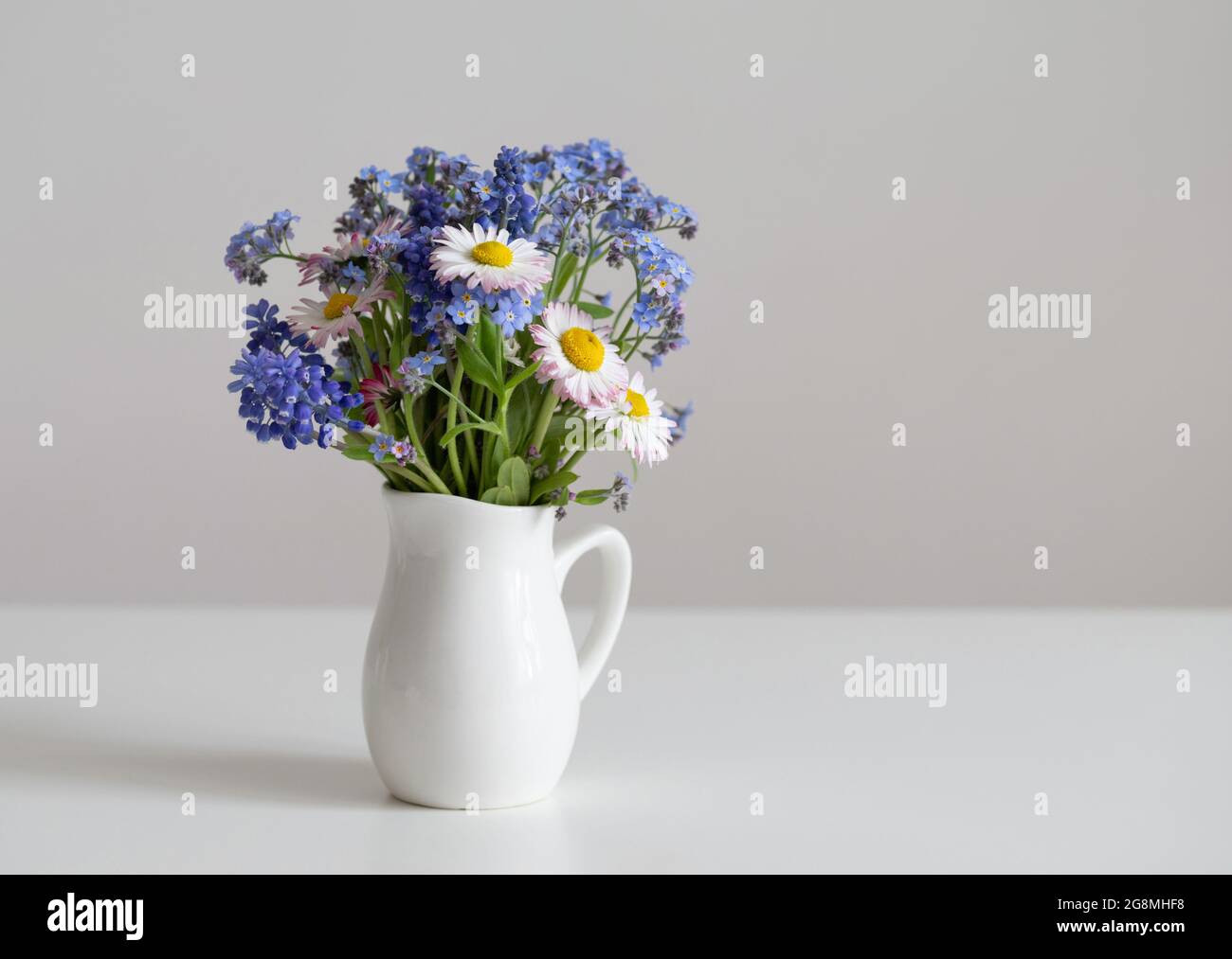 Flowers bouquet in a jug on a white table. Daisies and forget-me-nots ...