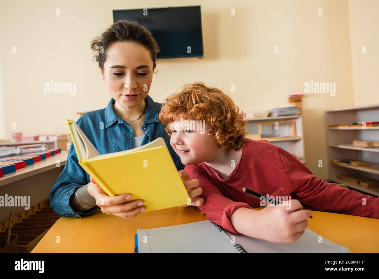 redhead boy holding pen and reading book in hands of young teacher ...