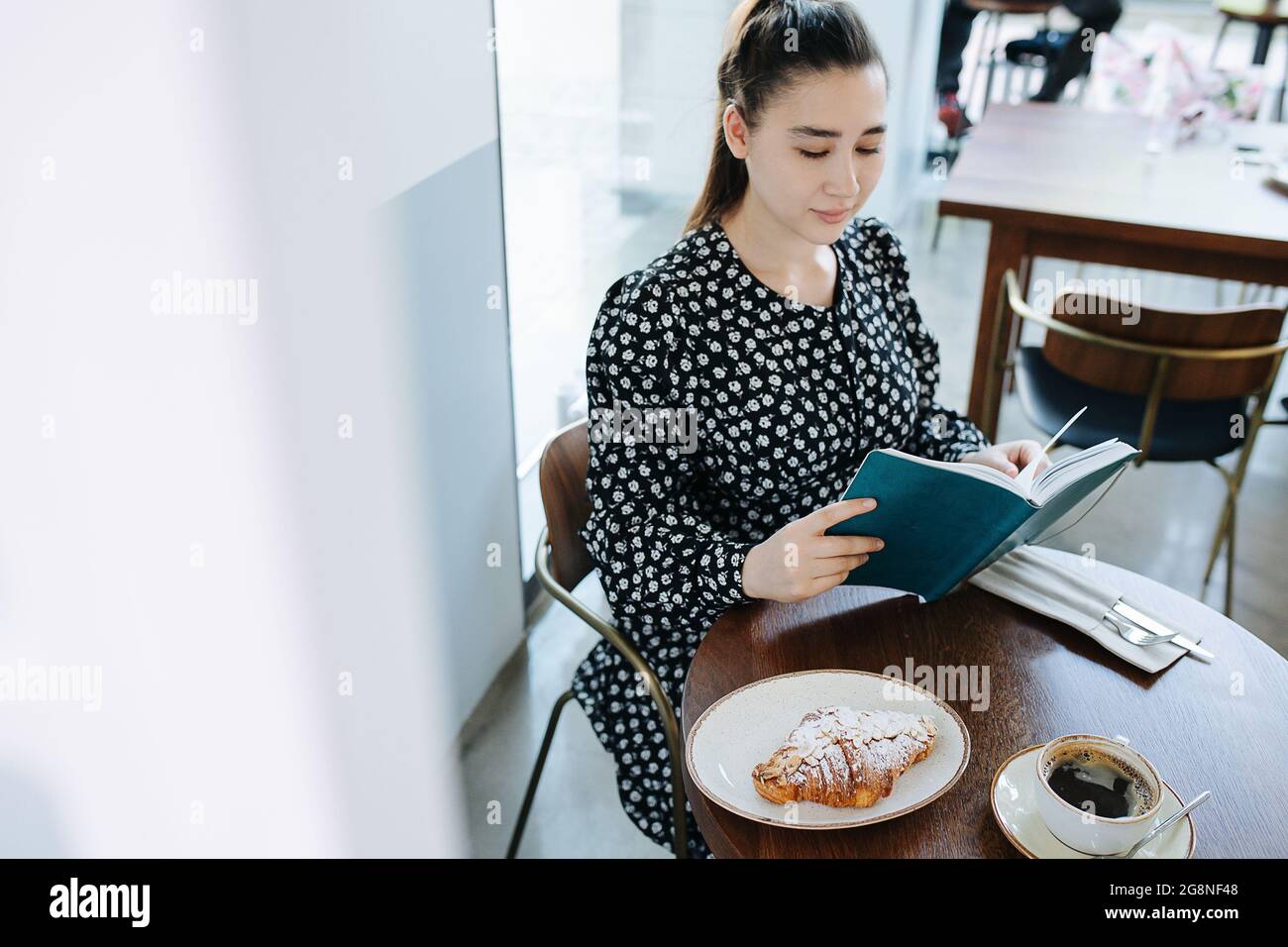 Pretty woman is reading a book in a cafe. Sitting behind a small round ...