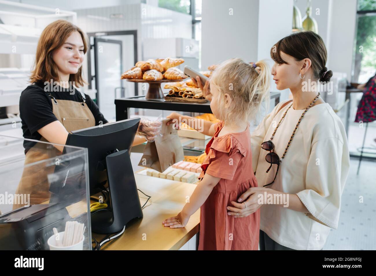 Friendly cashier handing pastry goods in a bag to mom with a daughter ...
