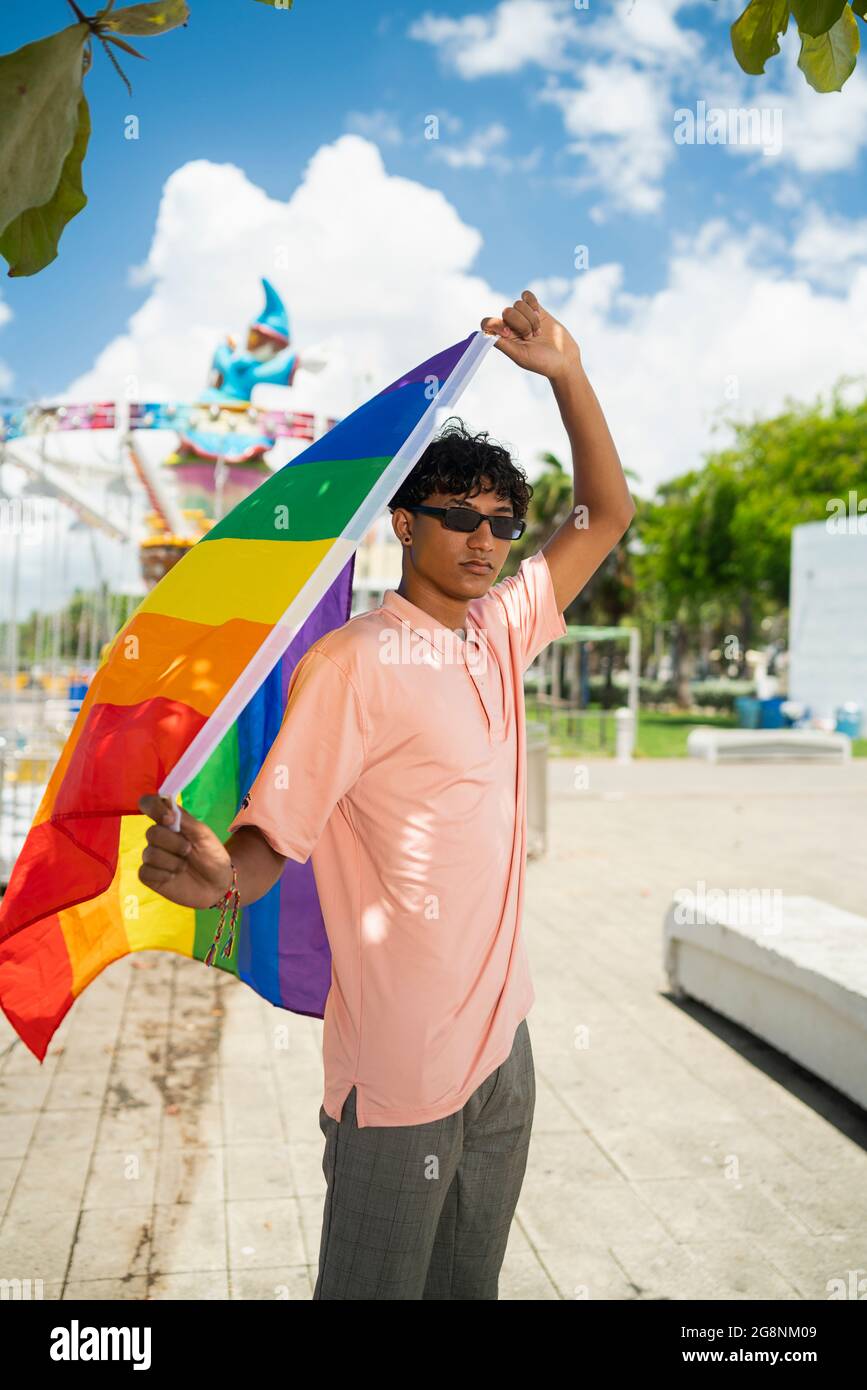 Portrait of young Hispanic gay boy looking at camera, holding LGBT flag ...