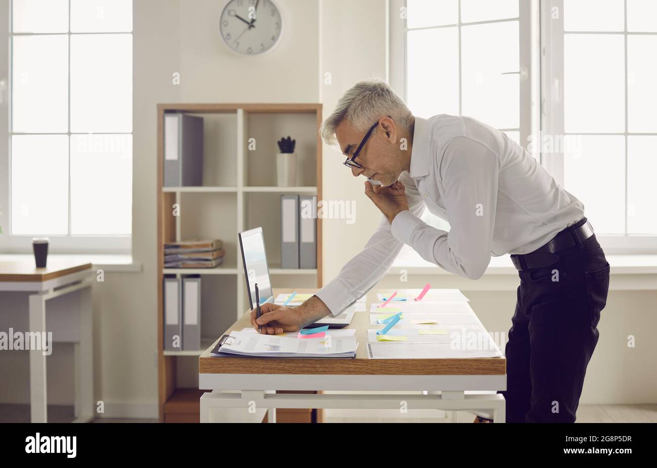 Serious senior man in the office writes notes while making a phone call ...