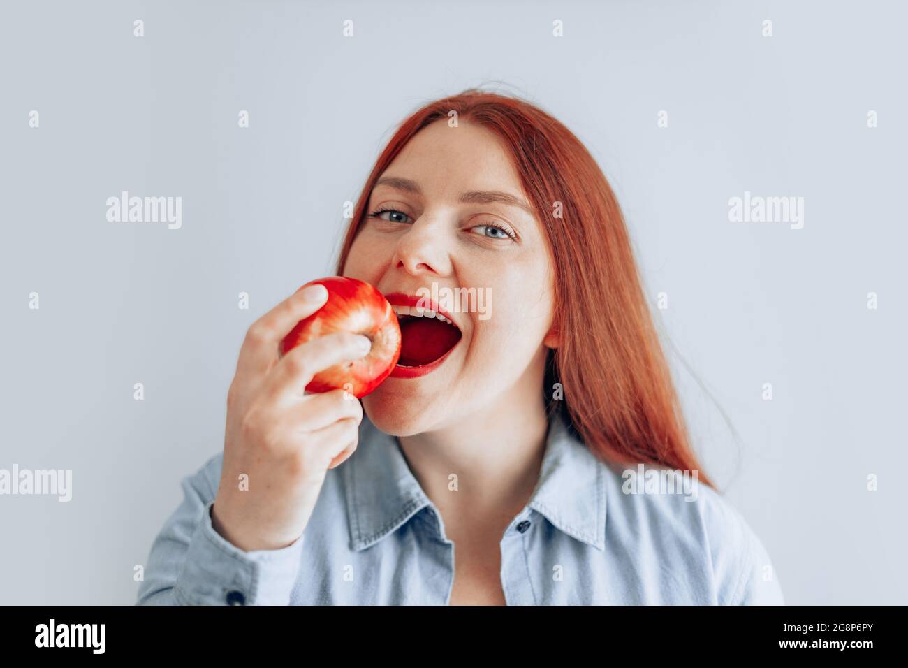 Portrait of a cheerful young woman eating red apple on gray wall ...