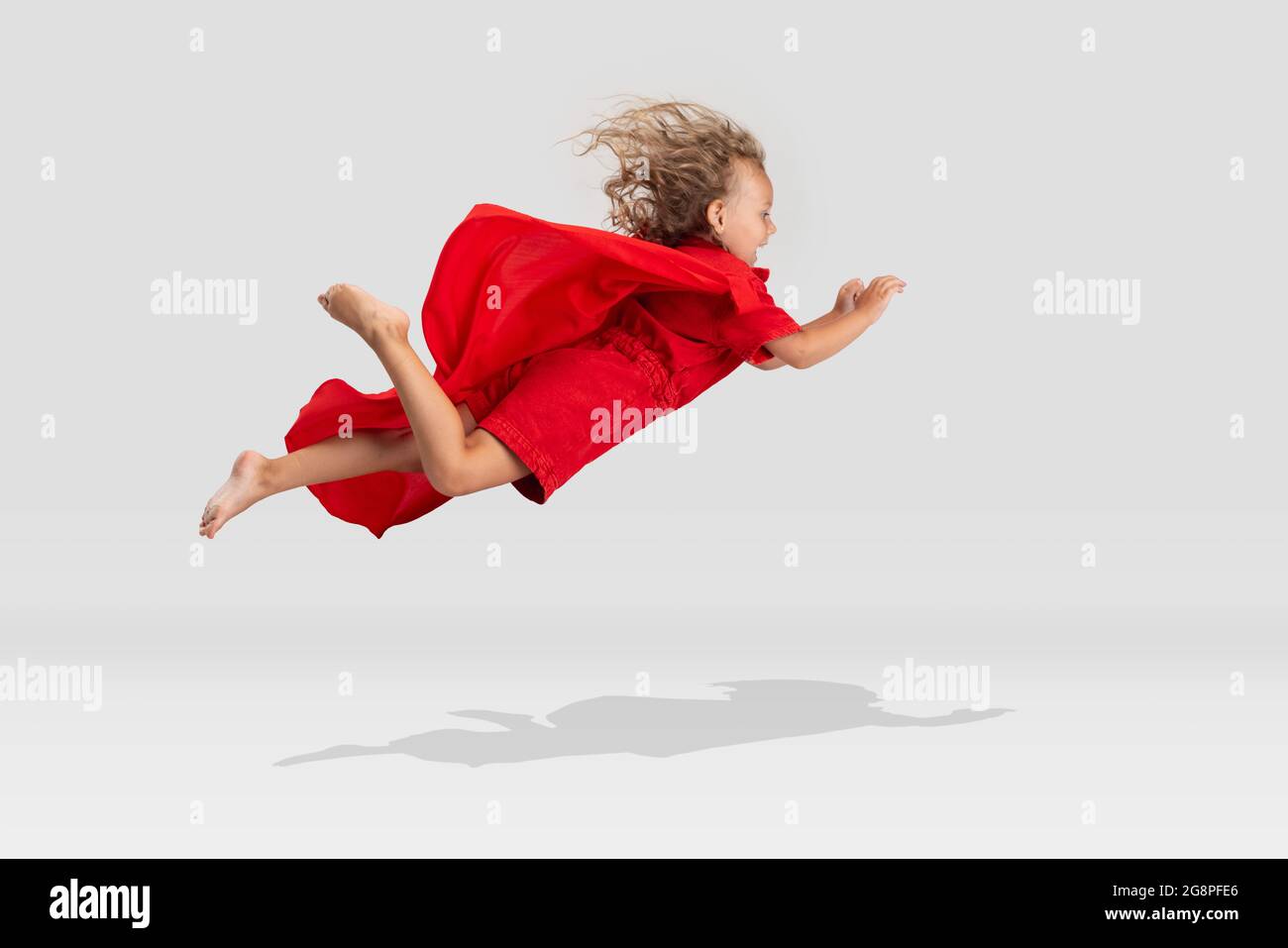 Flying above the ground. Cute little smiling girl in red cape isolated ...