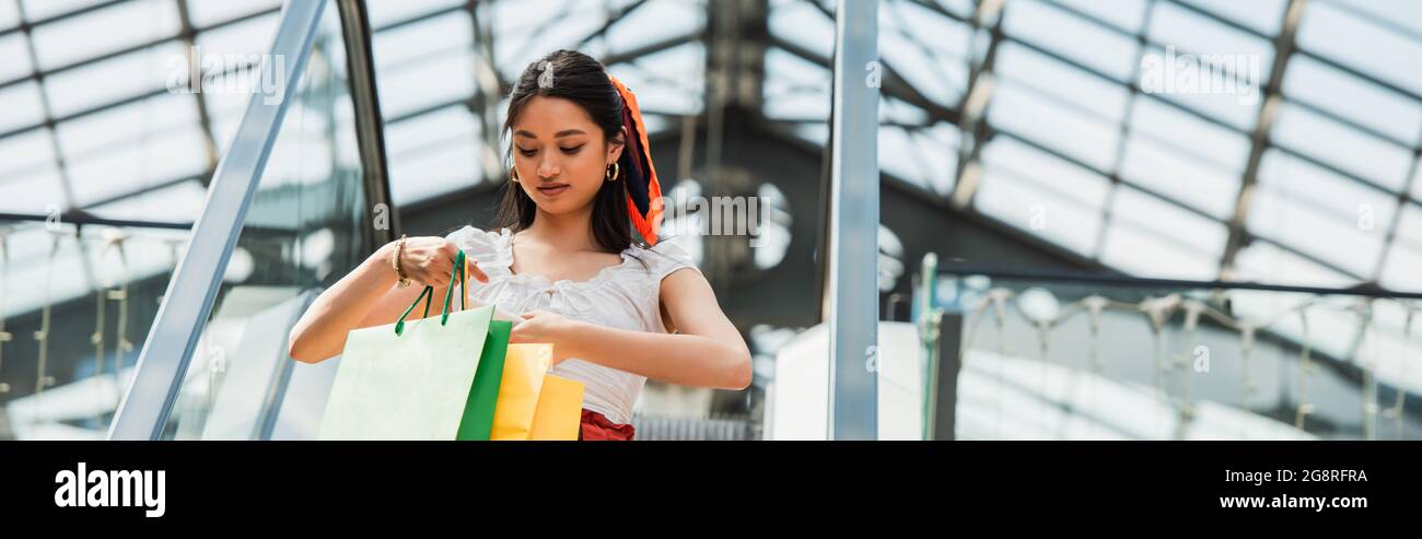 stylish asian woman looking into shopping bags on escalator, banner ...