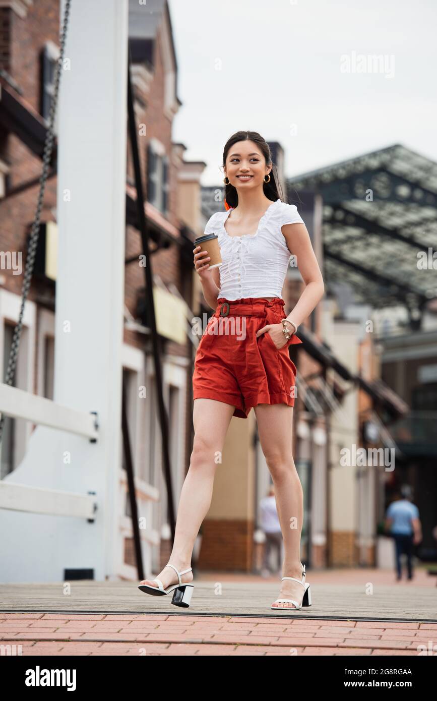 joyful asian woman holding coffee to go while walking on urban street ...