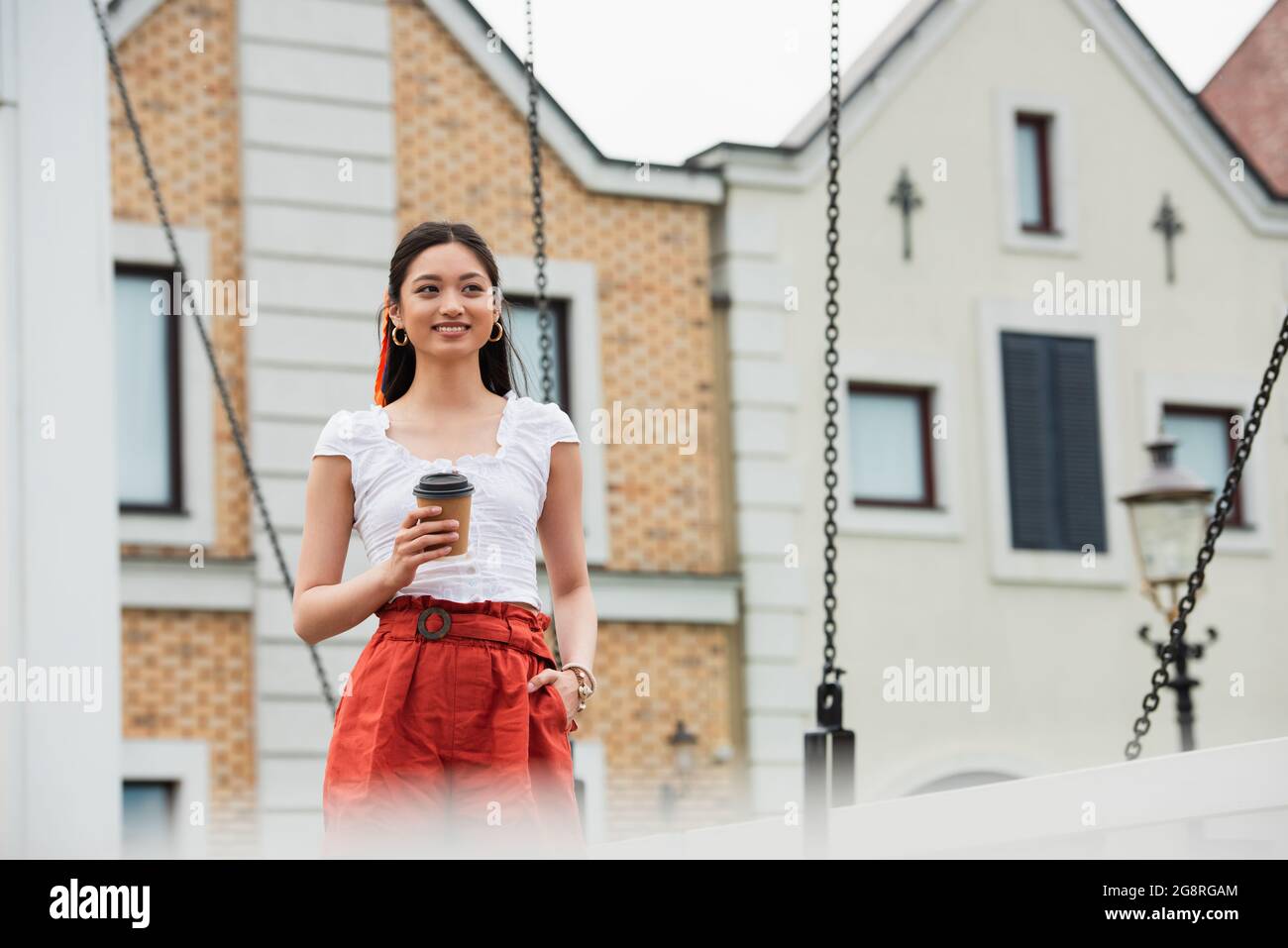 smiling asian woman with coffee to go standing with hand in pocket of ...