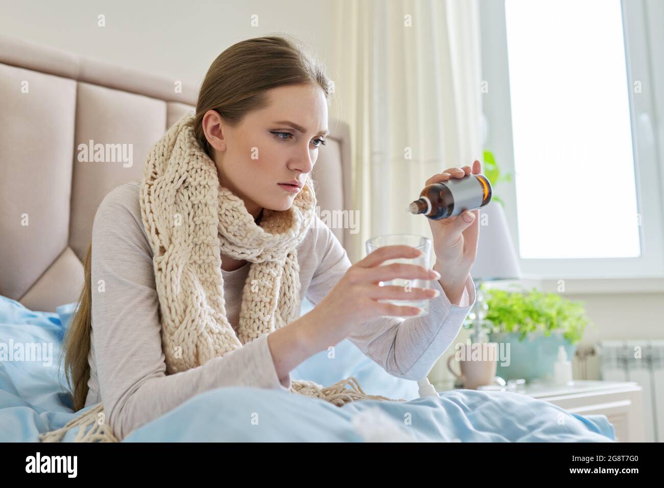 Sick woman at home in bed adding drops of medicine to a glass of water ...