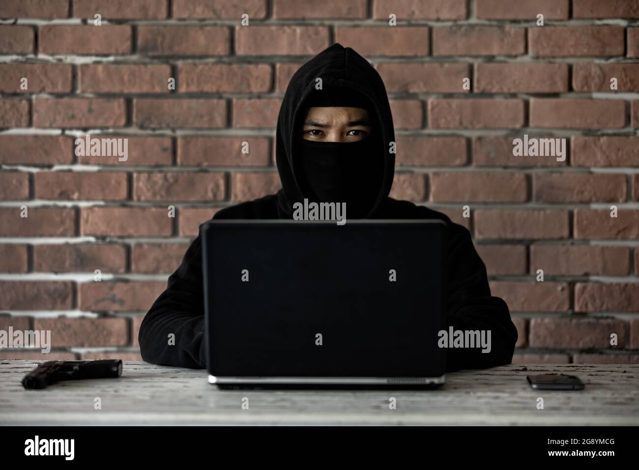 Cyber robber in black hood with notebook computer, gun and mobile phone ...