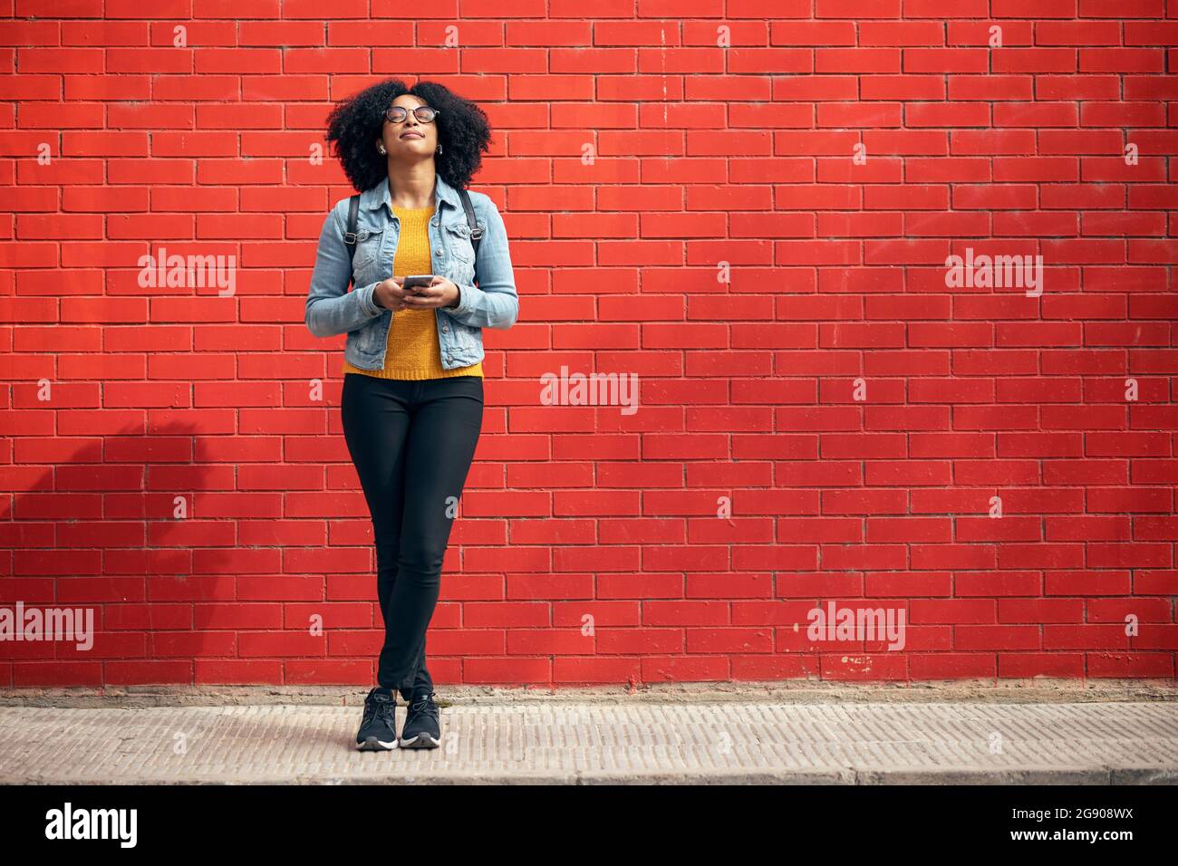 Woman looking up holding smart phone while standing near red brick wall ...