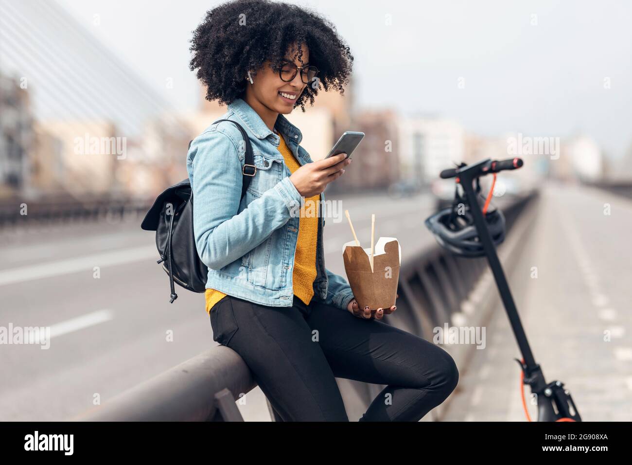 Smiling woman using smart phone holding food while leaning on railing ...
