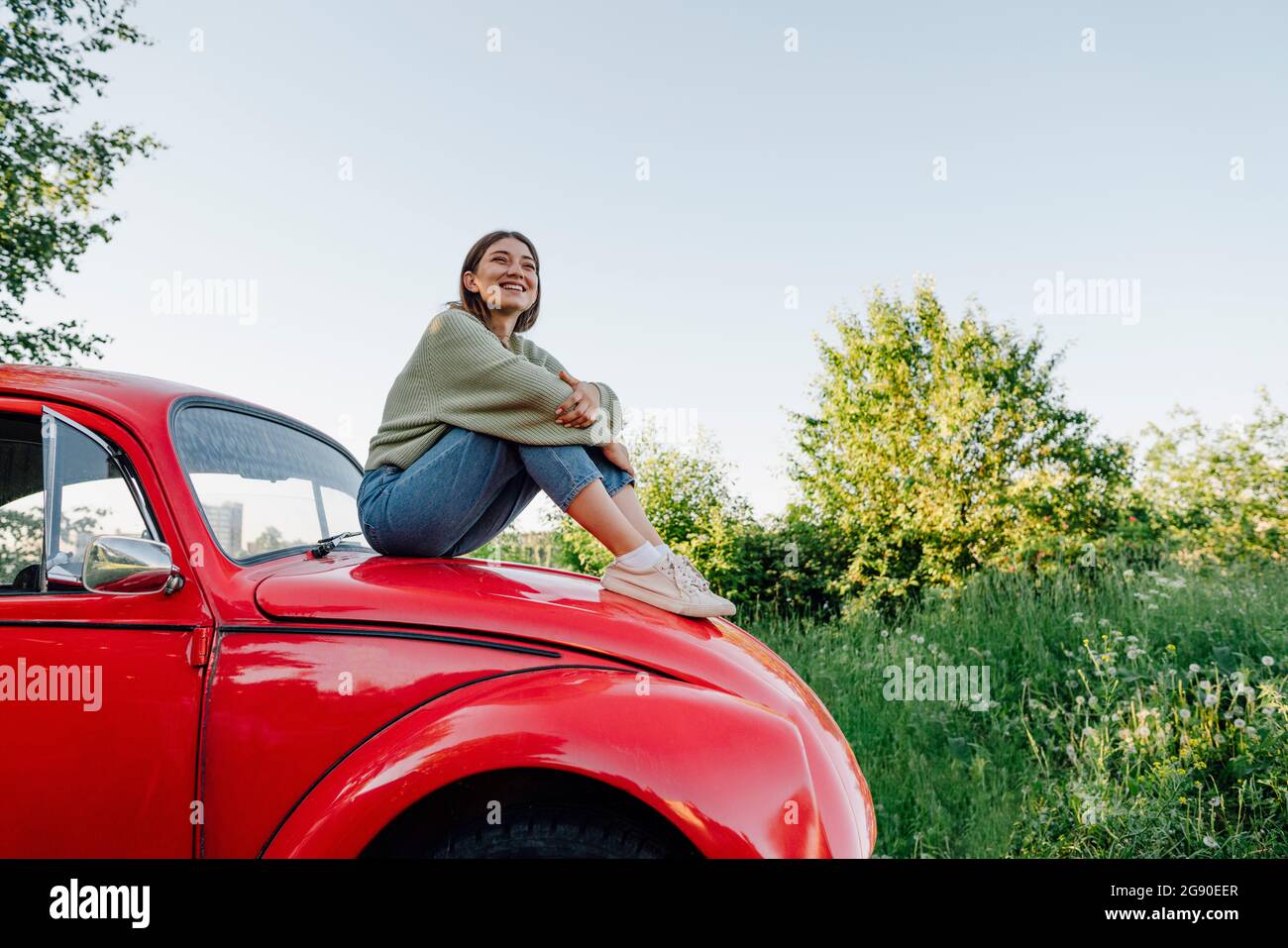 Smiling young woman hugging knees sitting on car hood Stock Photo - Alamy