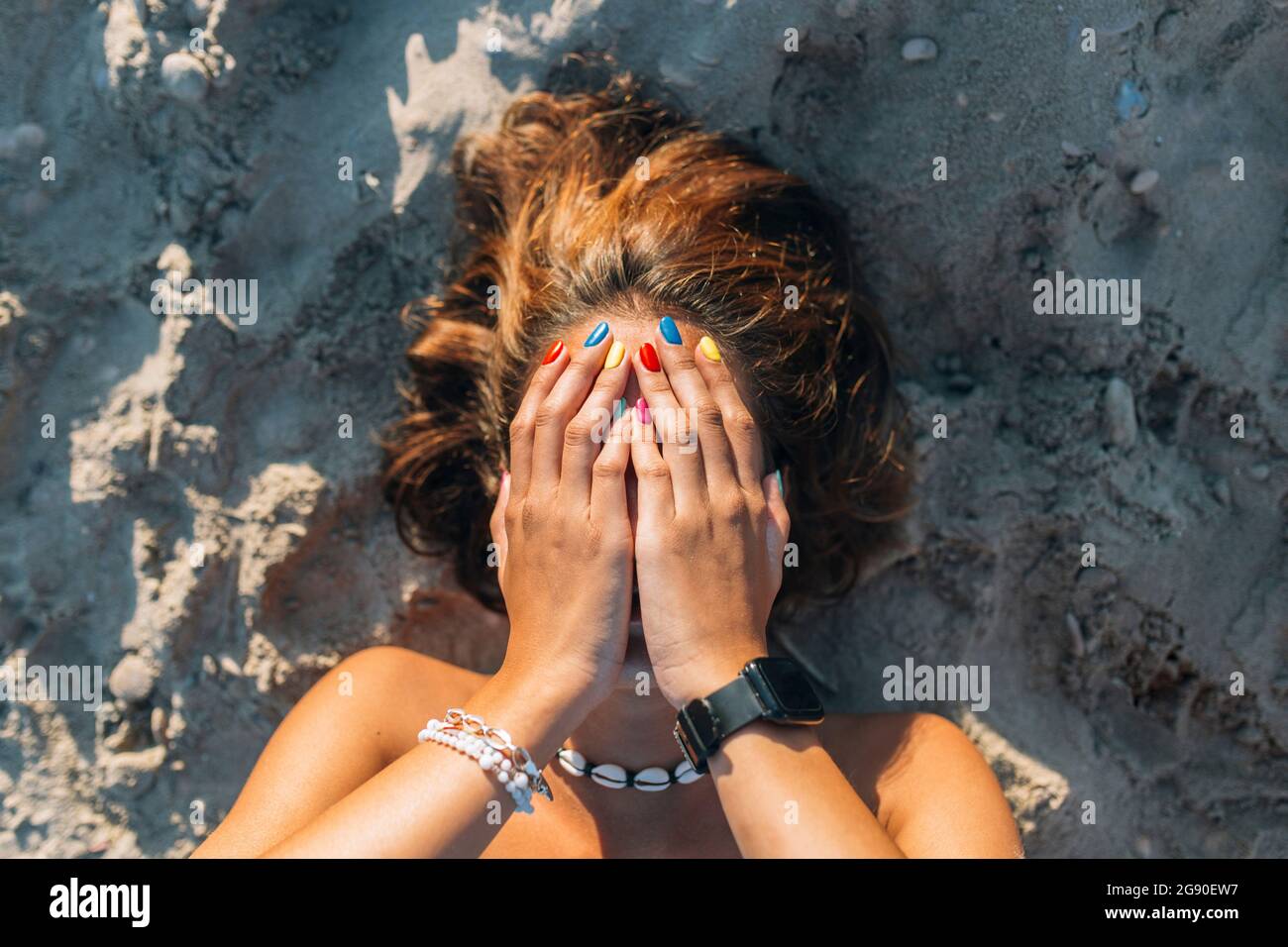 Woman covering face with hands while lying on sand during sunny day ...