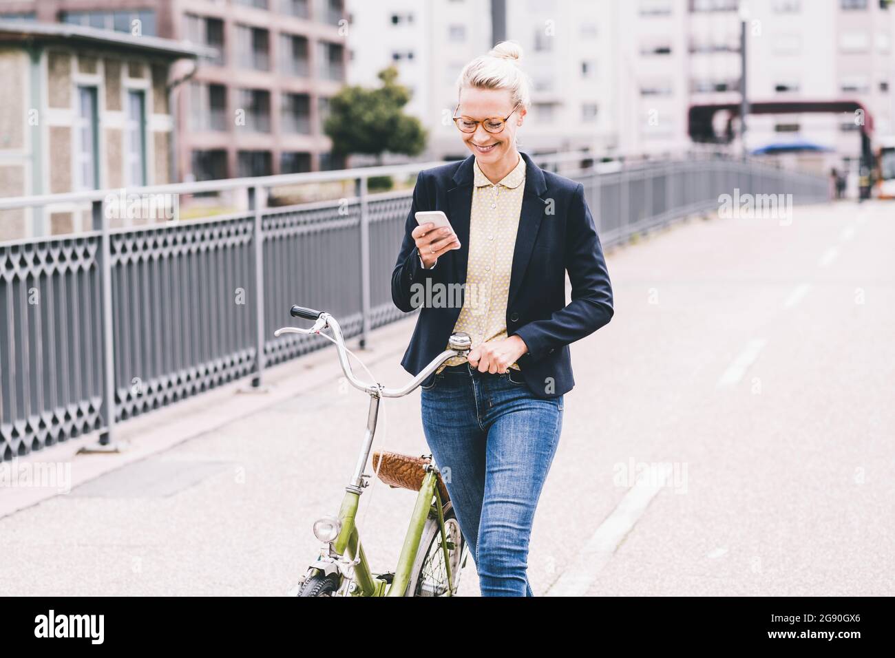 Blond female commuter wheeling bicycle while using mobile phone on ...