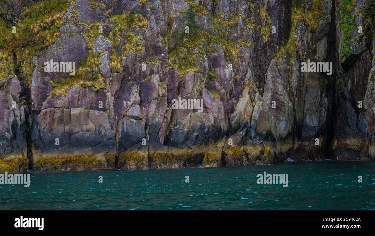 Cliff covered in greenery with colorful layers by the green water Stock ...