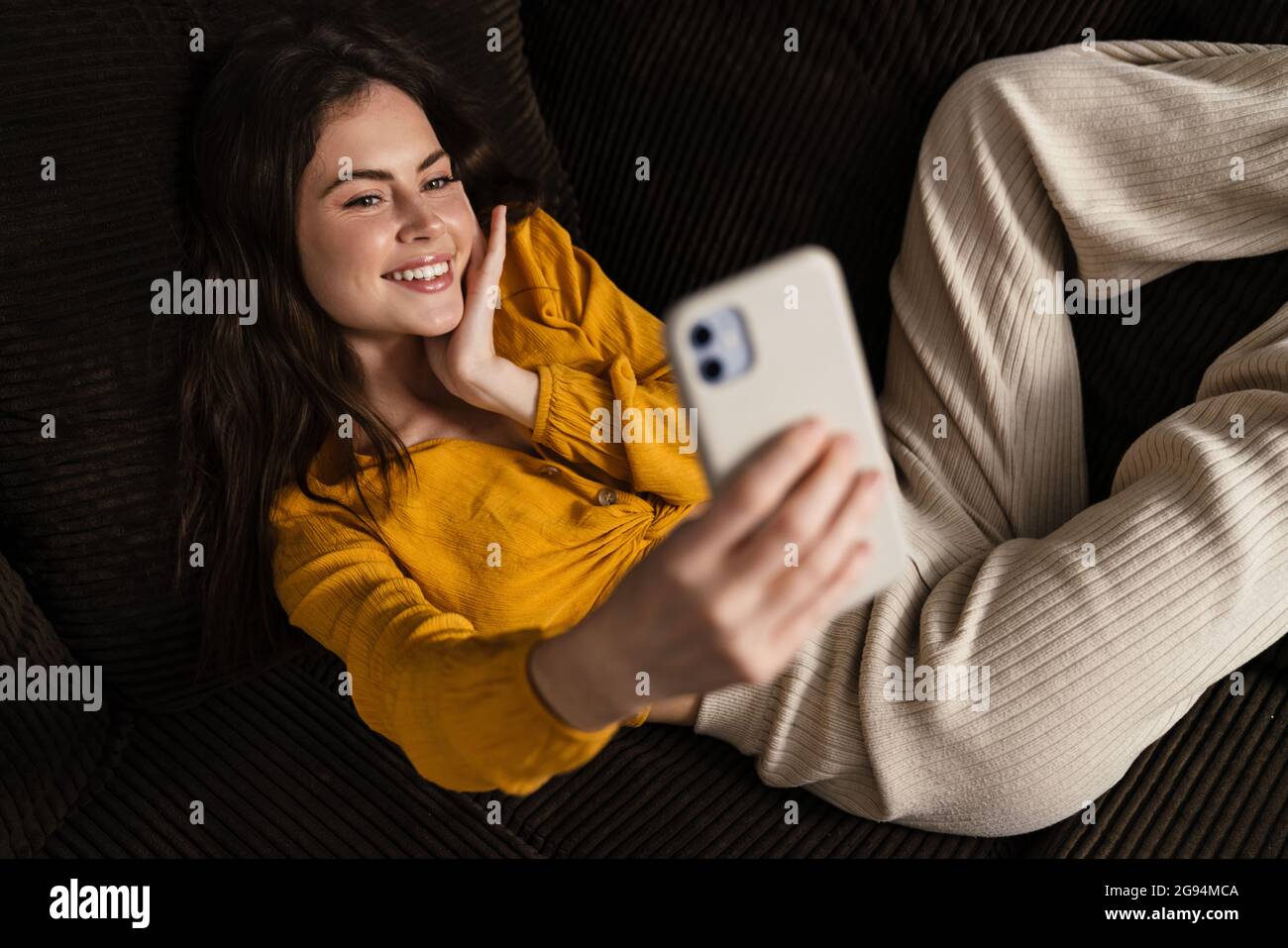 Smiling young brunette woman using mobile phone while laying on a couch ...