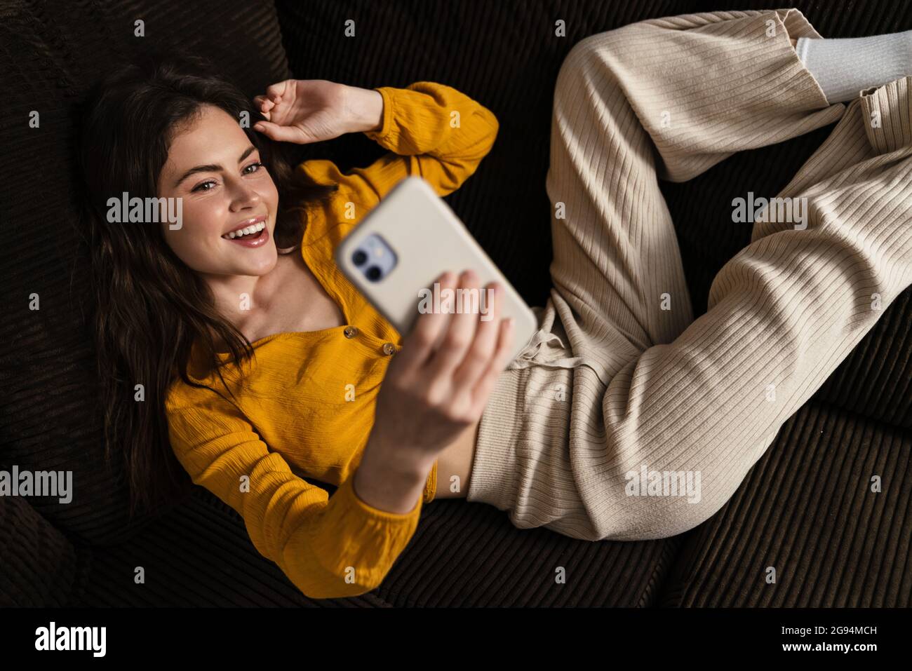 Smiling young brunette woman using mobile phone while laying on a couch ...