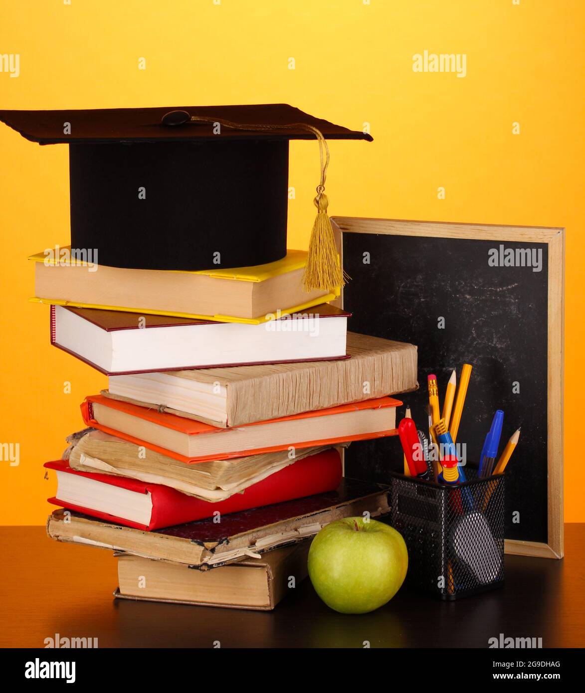 Books and magister cap against school board on wooden table on yellow ...