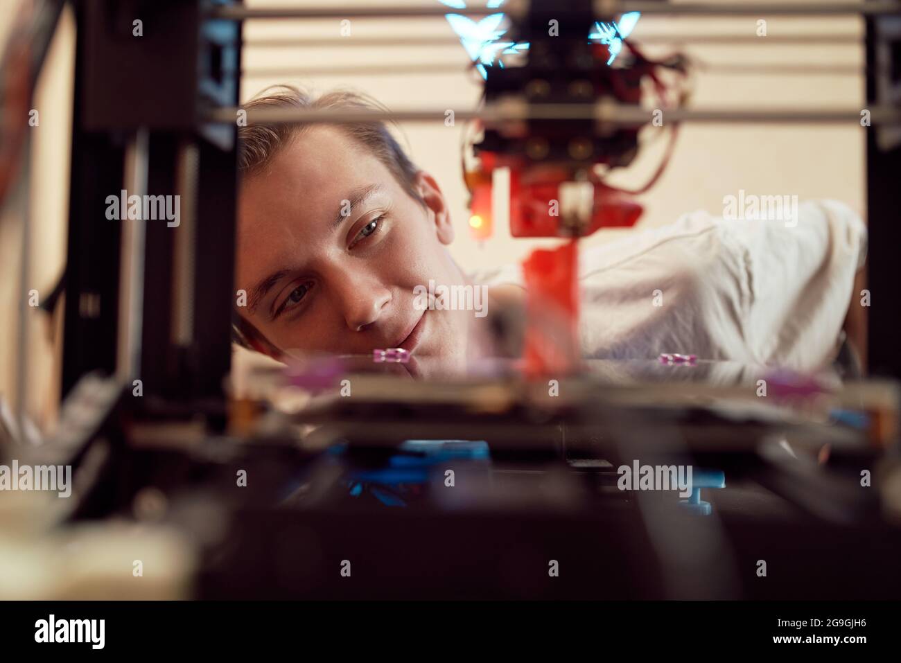 Young guy smiling and looking at 3D printer creating red plastic model ...