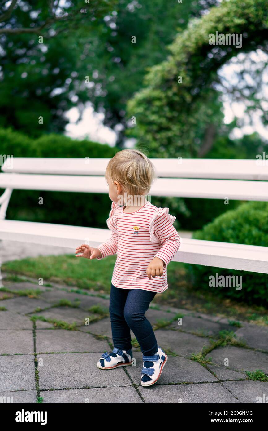 Little girl stands at a wooden bench in the park, turning her head back ...
