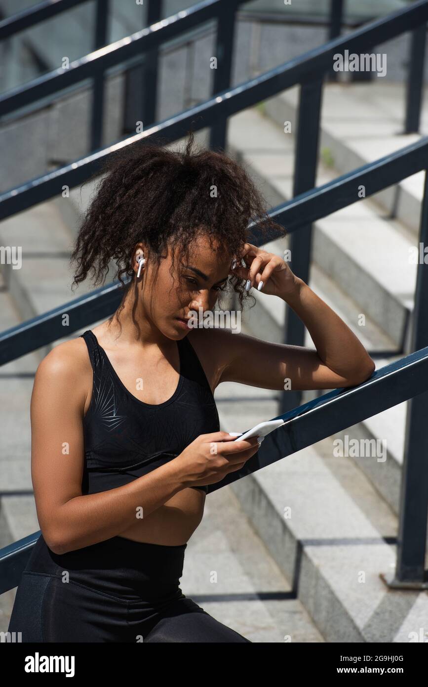 African american sportswoman in earphone using cellphone on stairs ...