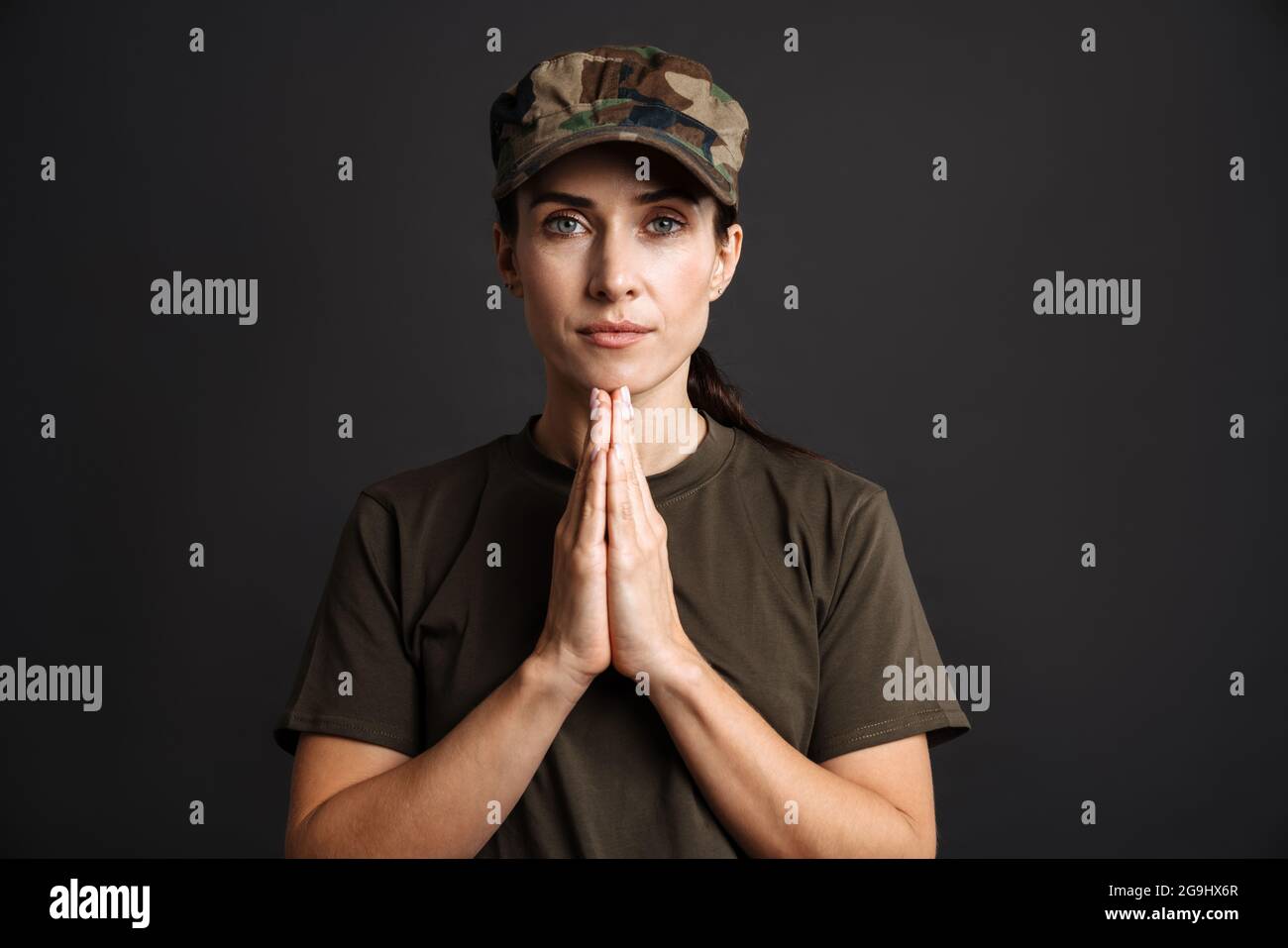 Calm beautiful soldier woman holding palms together isolated over black ...