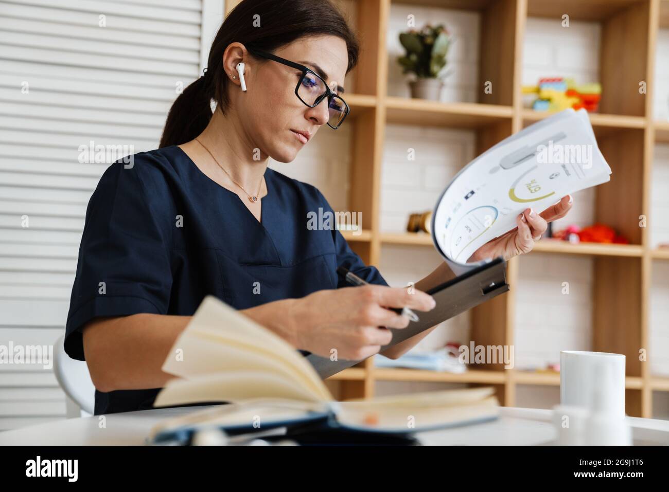 Focused woman doctor using earphone while working with papers at home ...