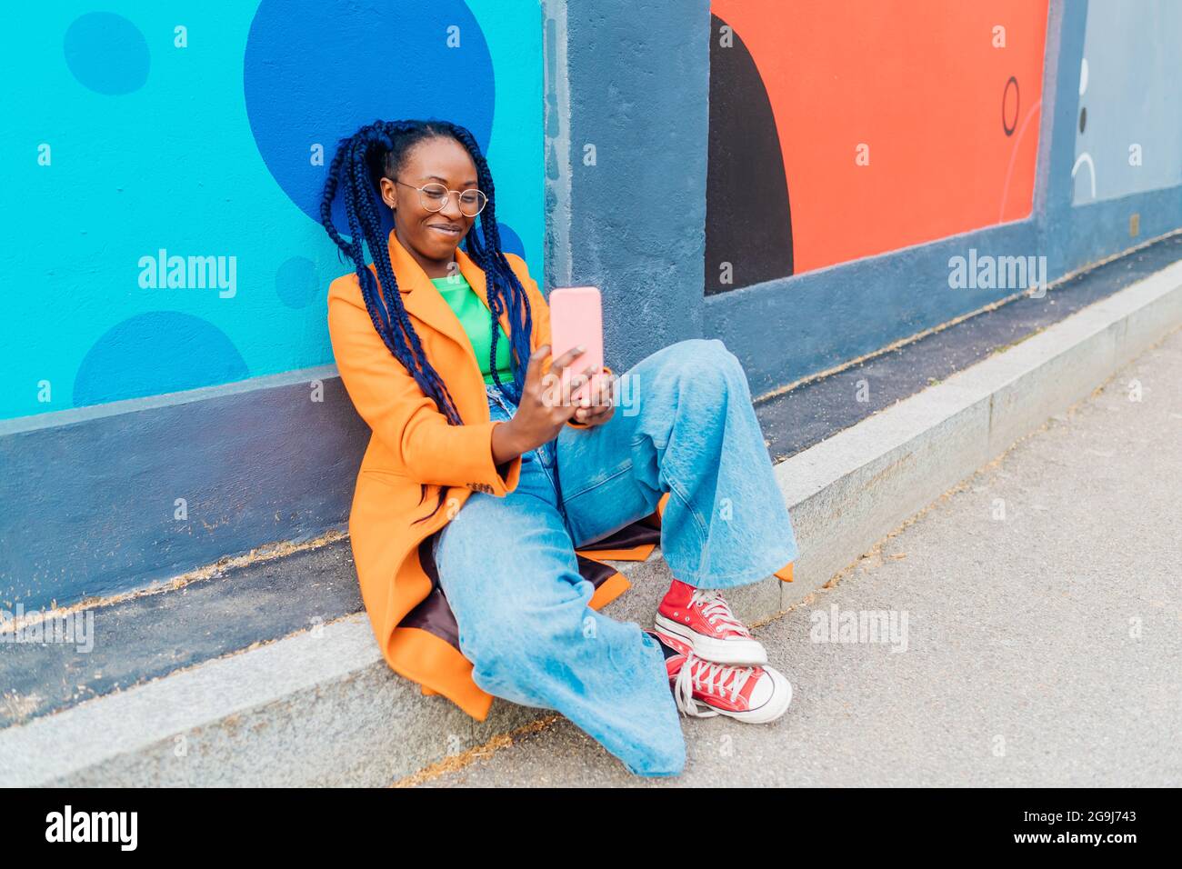 Italy, Milan, Woman with braids sitting by colorful wall, using smart ...