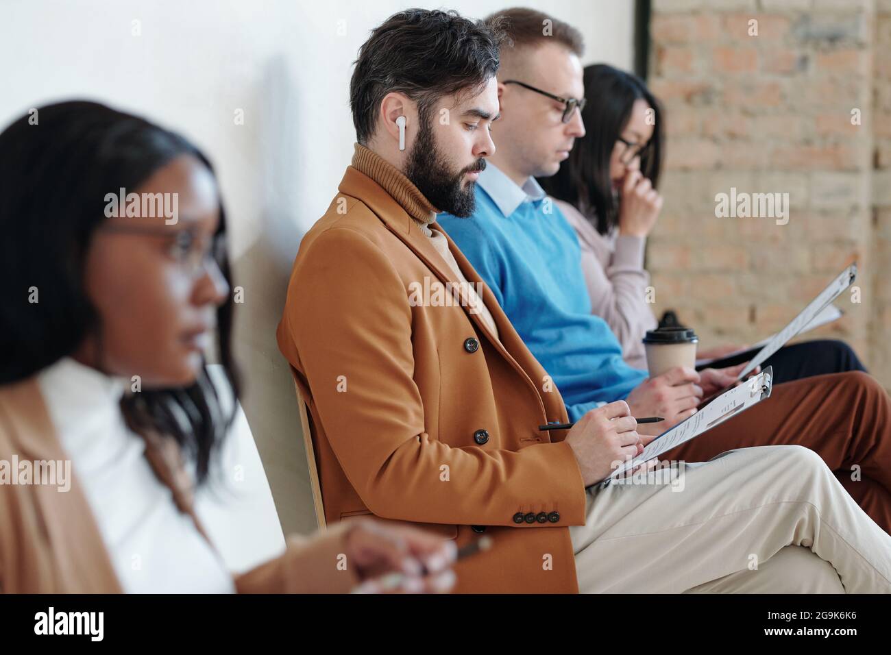 Group of serious young intercultural people sitting in row against wall ...