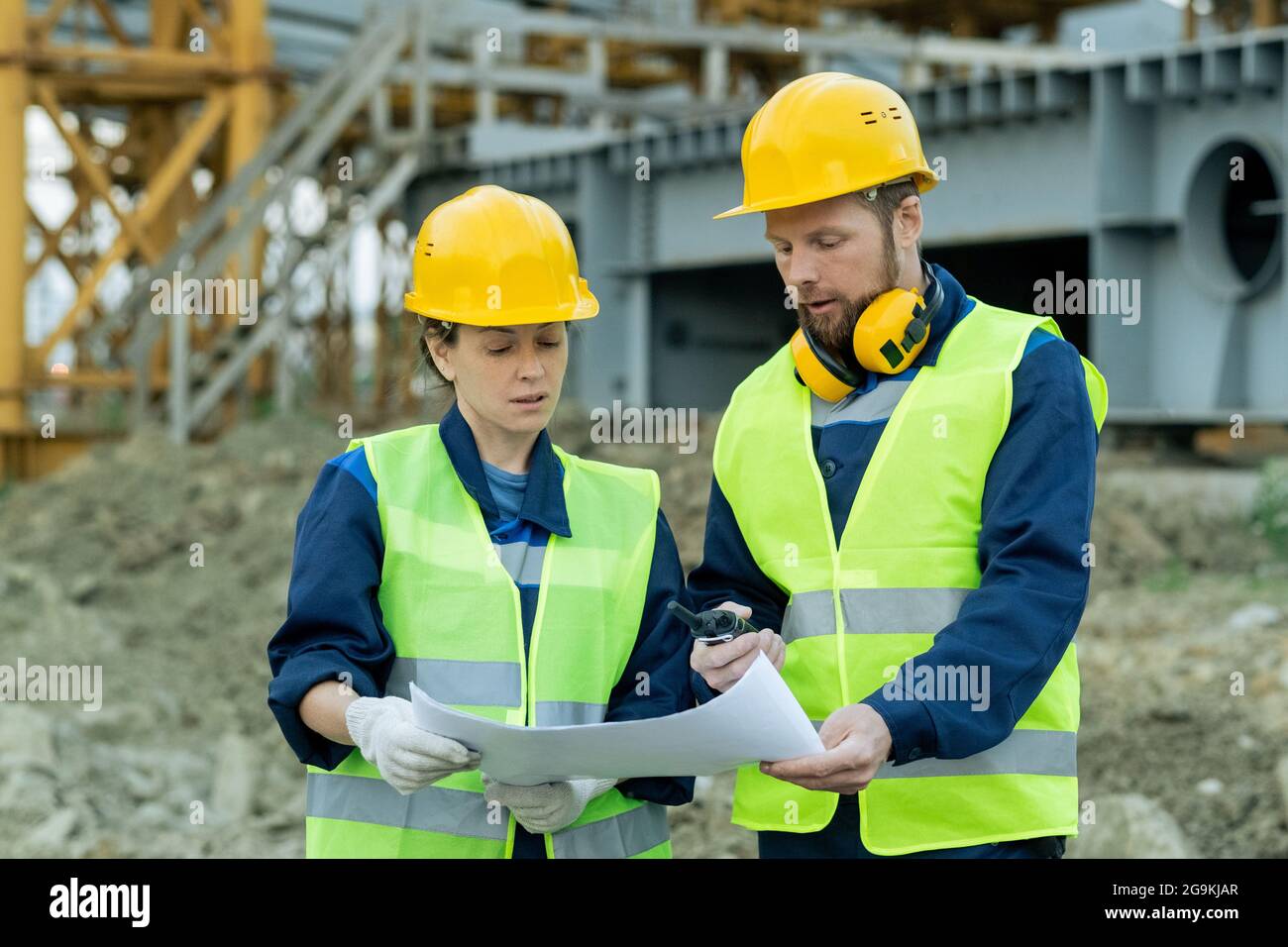Two engineers in workwear and in work helmets examining blueprint while ...