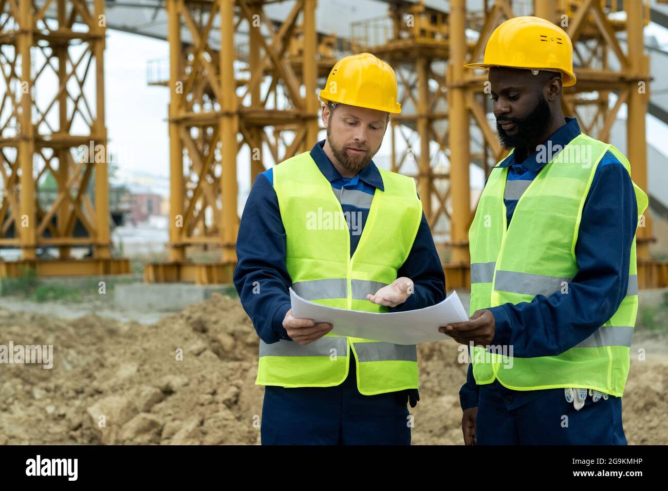 Two engineers in reflective clothing examining blueprint and discussing ...