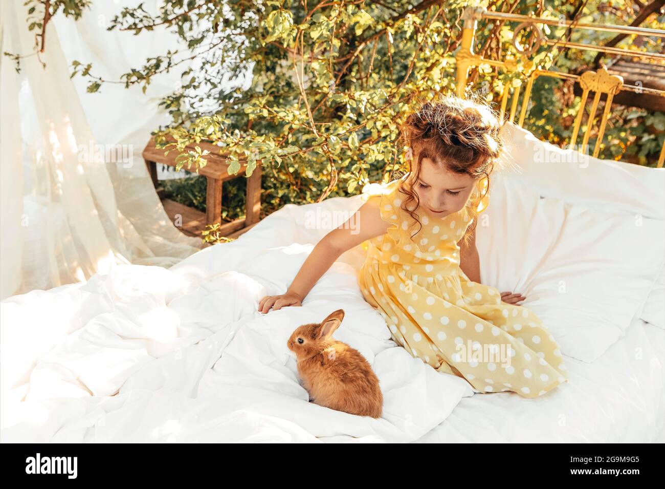 Little pretty girl in summer dress sitting outside on the bed with ...