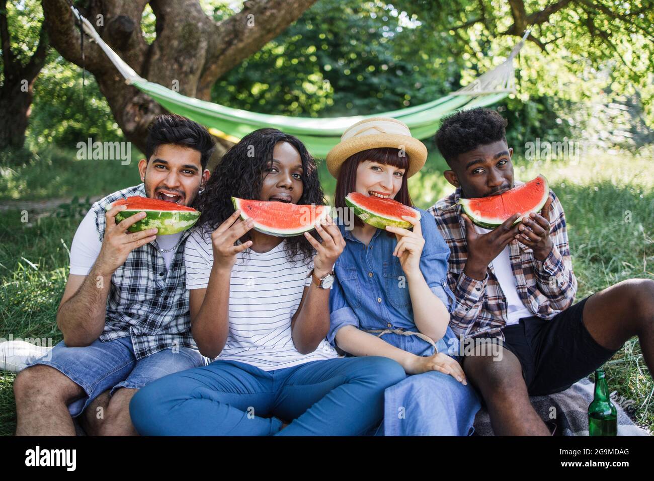 Group of four multicultural people eating sweet ripe watermelon while having picnic at green ...