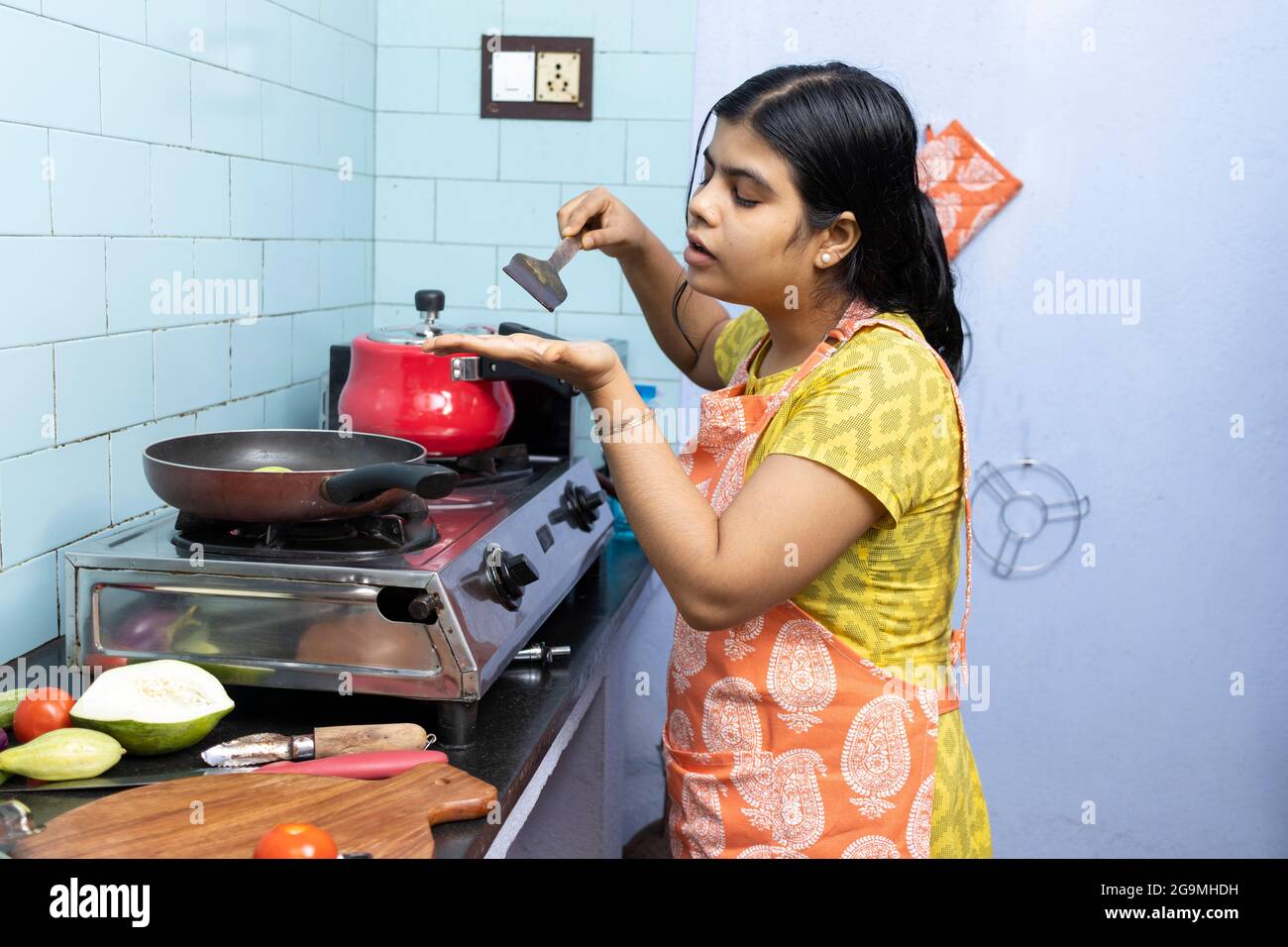 A Pretty Indian Young Woman Wearing Apron Cooking And Tasting Food In